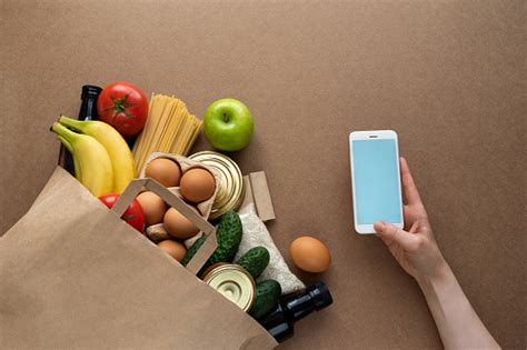 음식용품이 담긴 종이 봉투와 중성 베이지색 배경에 전화로 여성손 음식 배달 검역 기부 코로나 바이러스 개념에 대한 모바일 주문