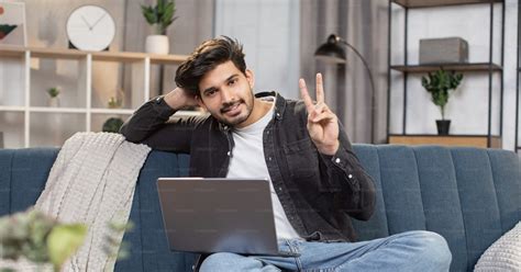집에서 모바일 장치를 사용하는 젊은이들의 개념 웃는 수염을 기른 아라비아 인도 남자 소파에 앉아 집에서 노트북 컴퓨터로 작업하며 카메라에 승리의 신호를 보여줍니다 사진