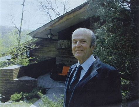 A Man In A Suit And Tie Standing Next To A Cabin On The Side Of A Hill