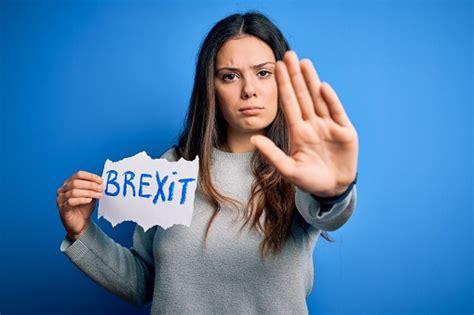 진지하고 자신감있는 표정 방어 제스처와 정지 기호를하고 열린 손으로 파란색 배경위에 브렉시트 메시지와 함께 종이를 들고 젊은 아름다운 갈색 머리 여자 Referendum에