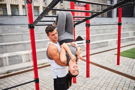 쾌활한 날씬한 여성이 야외에서 크로스바에 위쪽으로 매달려 있고 근육질의 남자가 그녀를 돕고 있다 사진 Unsplash의 운동 이미지