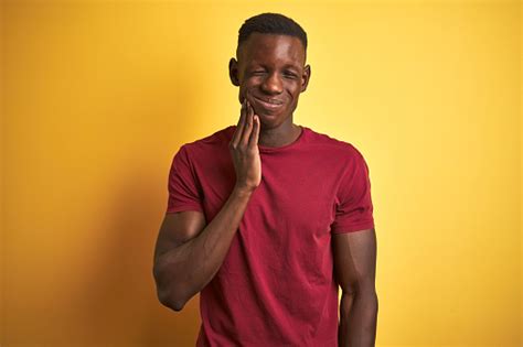 치아에 치통이나 치과 질환으로 인해 고통스러운 표정으로 손을 만지는 고립 된 노란색 배경 위에 서있는 빨간 티셔츠를 입은 젊은 아프리카 계 미국인 남자 치과 의사 개념 T