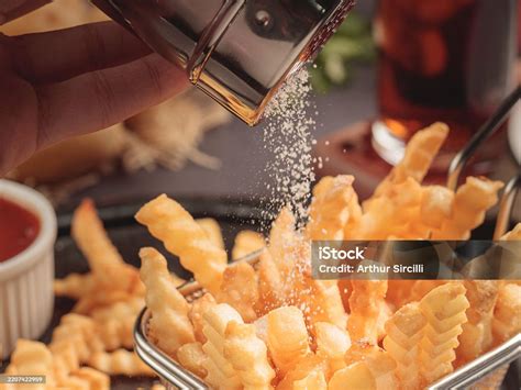 금속 셰이커의 소금을 크링클 컷 감자 튀김 바구니에 손으로 흔듭니다 빨간 소스가 담긴 작은 흰색 그릇과 어두운 음료 한 잔이 흐릿한 배경에 보입니다 Comfort Food에