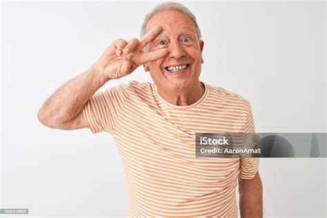 고립 된 흰색 배경 위에 서있는 줄무늬 티셔츠를 입은 선임 회색 머리의 남자가 얼굴에 손가락으로 평화 기호를하고 승리를 보여주는