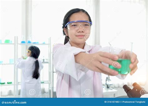 Cute Young Scientist Schoolgirl Wears Lab Coat And Safety Glasses Holds Green Chemical Beaker