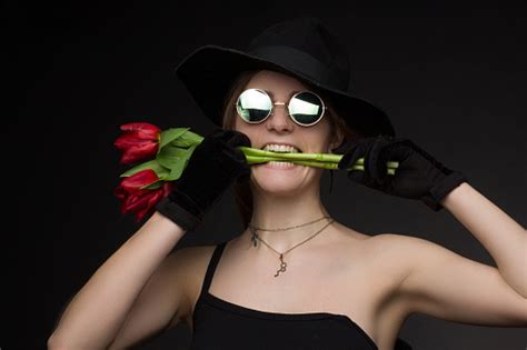 모자와 선글라스를 입은 젊은 여성의 스튜디오 초상화가 그녀의 입에 빨간 튤립 꽃다발을 들고 검은 색 배경에 검은색에 대한 스톡 사진 및 기타 이미지 Istock