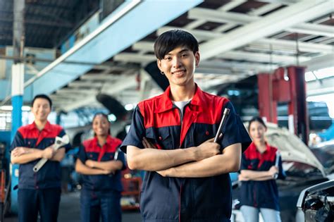 아시아 자동차 엔지니어 사람들은 정비사 차고에서 헬멧 작업을 착용합니다제복을 입은 젊은 자동차 정비사는 카메라를 보고 웃으며 차를 검사하고 있습니다 자동차 수리 공장에 대한