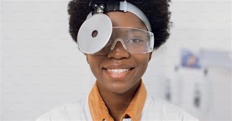 젊은 친절한 미소를 짓고 있는 아프리카계 미국인 여성 의사 이비인후과 의사 Hed 정면 반사경과 보호 안경을 착용하고 현대적인 조명 사무실에서 카메라를 향해 포즈를 취하는