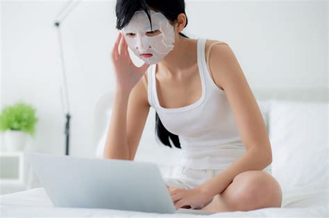 침실에서 침대에 노트북 컴퓨터에 앉아 시트 얼굴 마스크를 가진 아름다운 젊은 아시아 여성 집에서 주름의 스킨 케어에 대한 얼굴 마스크를 적용 뷰티 걸 피부 관리 건강 및