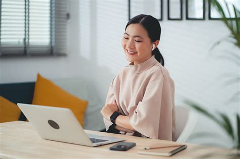 이어버드를 착용한 아시아 여성은 홈 오피스에 앉아 화상 회의에서 노트북을 사용하여 온라인 작업을 즐깁니다 Brand Name Smart Phone에 대한 스톡 사진 및 기타