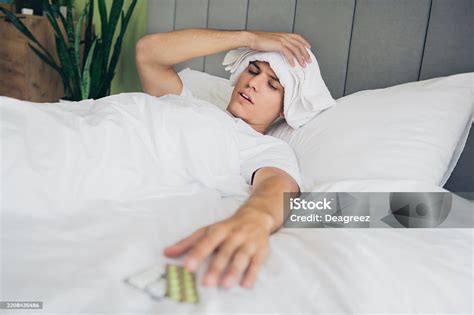 머리에 수건을 쓰고 침대에서 쉬고 있는 젊은이가 휴식과 가정의 편안함을 보여주는 약을 먹기 위해 손을 뻗습니다 20 24세에 대한 스톡 사진 및 기타 이미지 Istock