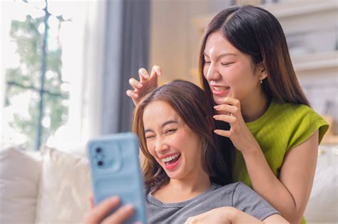 아시아 친구 소파에서 카메라 폰으로 자화상을 이야기하는 젊은 여성 행복한 젊은 여성이 여자 친구에게 스마트 폰으로 소셜 미디어 콘텐츠를 보여줍니다 그들은 집 소파에 앉아