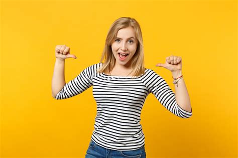 카메라를 찾고 줄무늬 옷에 쾌활 한 젊은 여자 스튜디오에서 노란색 오렌지 벽 배경에 고립 자신에 엄지손가락을 가리키는 사람들은 진심으로 감정 라이프 스타일 개념입니다 복사