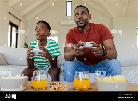 African American Father And Son Gripping Controllers On Sofa At Home