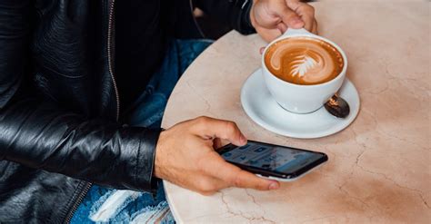 ~을 사용하여 검은색 가죽 자켓 도자기 컵 라떼 아트 라운드 테이블 사람 스마트폰 잡고 있는 커피 한 잔 하이앵글 샷에 관한 무료 스톡 사진