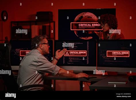 Mature Programmer Sitting In Front Of Computer Monitors With Critical