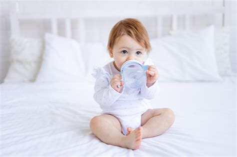 흰 옷을 입고 침대에 누워 6개월 된 여자 아기가 물이나 우유 한 병을 빨고 집에서 면 침대에 누워 아침에 일어나 먹는 어린 아이 이유식 개념 6 11 개월에 대한 스톡