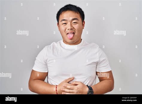 Young Chinese Man Standing Over White Background Sticking Tongue Out