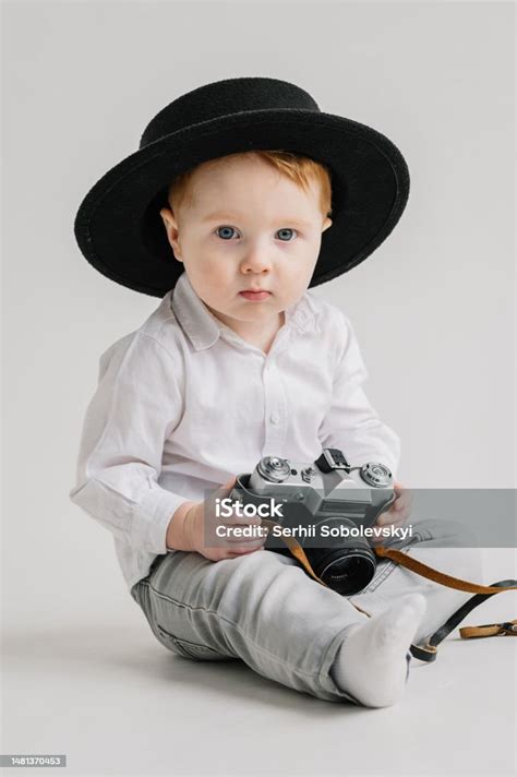 재미있는 아이는 흰 벽에 고립된 복고풍 빈티지 카메라를 들고 있다 모자를 쓴 아이가 바닥에 앉아 사진 카메라를 가지고 놀고 있다