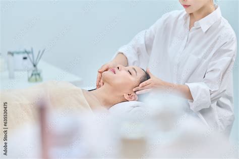 아시아 한국의 젊은 여성이 스파 또는 피부관리전문점 또는 피부과 병원에서 침대에 누워 베이지색 대형 타올을 덮고 눈을 감고있고 하얀색 셔츠를 입은 전문관리사가 손으로 얼굴