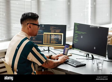Side View Of Asian It Developer Typing On Keyboard With Programming Code On Computer Screen