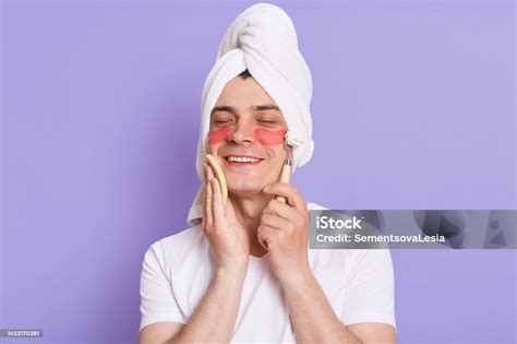 수건을 입은 즐거운 청년의 실내는 눈 주름 방지 패치를 사용하여 손에 스폰지를 들고 롤러로 얼굴 마사지를하고 아침 미용 절차를 즐깁니다 35 39세에 대한 스톡 사진 및
