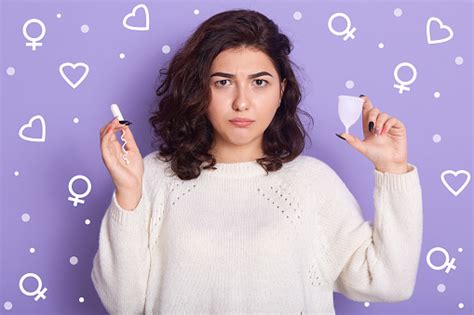 만족하지 혼란 갈색 머리의 실내 스튜디오 샷은 카메라를 직접 보고 두 손에 위생 패드와 생리 컵을 들고 라일락 배경 위에 고립 된 서 생리 개념 개념에 대한 스톡 사진 및