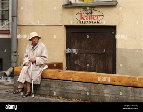 An Old Lady Sitting On A Wall With A Walking Stick Outside An Abandoned