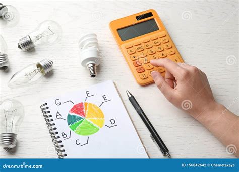 Woman With Calculator Energy Efficiency Rating Chart And Light Bulbs At Table Stock Photo