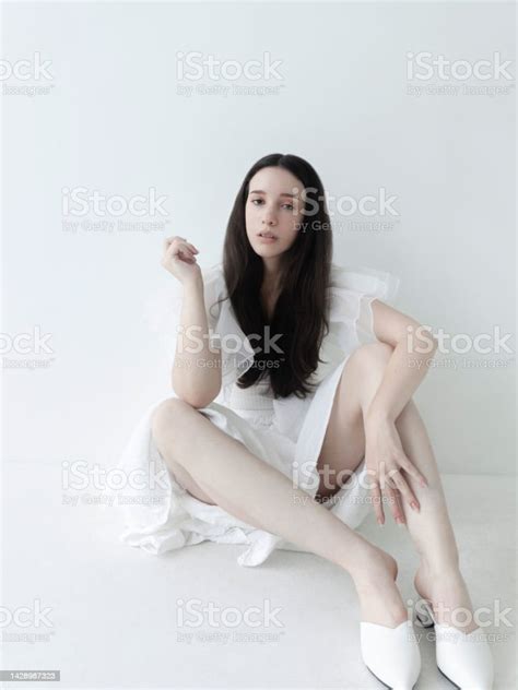 몽환적인 흰색 드레스와 신발로 바닥에 앉아있는 아름다운 젊은 갈색 머리 여자 전체 길이 바디 밝은 배경에 패션 모델 스튜디오