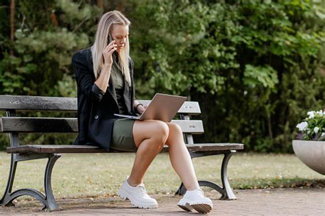 검은 블레이저를 입은 금발의 사업가가 여름 공원의 벤치에 앉아 노트북 컴퓨터에 타이핑하고 전화 통화를하고 몸짓을하고 야외에서 공부하고 있습니다 원격으로 일하는 여성 35