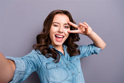 좋은 귀여운 명랑 사랑 스러운 매력적인 아름 다운 갈색 머리 여자 캐주얼 데님 셔츠 열린된 입 물결 모양 머리를 가진 눈 회색 배경 위에 절연 근처 Vsign를 보여주는의