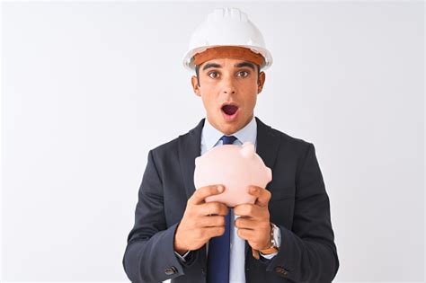 고립 된 흰색 배경 위에 돼지 저금통을 들고 헬멧을 쓰고 있는 잘생긴 건축가 남자는 깜짝 얼굴로 충격에 무서워하고 두려움과