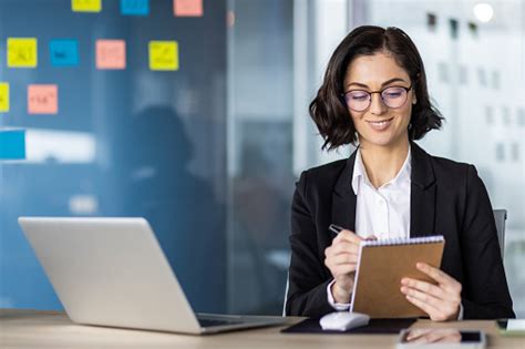 노트북 앞에 앉아서 메모를 하는 현대적인 사무실 환경에서 자신감 넘치는 사업가 전문적인 옷차림 그녀의 일에 집중하고 참여하며 다채로운 알림으로 둘러싸여 있습니다 개발에 대한