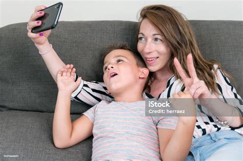 쾌활한 어머니는 승리 신호를 보여주는 작은 아들과 셀카를 만드는 스마트 폰을 들고 있습니다 엄마와 아이 소년은 집에서 함께 시간을 보내는 자화상을 찍는 재미를 가지고