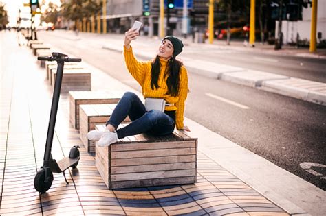 옆에 주차 된 전기 스쿠터와 대도시 지역 거리 보도에 벤치에 앉아 있는 동안 휴대 전화와 Selfie를 복용 하는 현대 여자