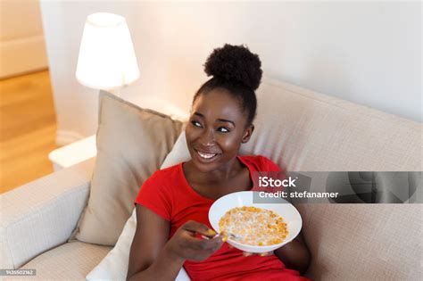 아침에 집에서 건강한 아침 식사를 먹고 거실에서 소파에서 휴식을 취하는 미소 짓는 여성 아름다운 행복한 아프리카 계 미국인 소녀 즐기는 음식 에 그릇 앉아 소파 아침 식사에