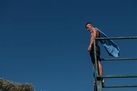 강 근처의 어린 십대 소년이 여름 파티에서 친구들과 즐거운 시간을 보내고 슈퍼 히어로 스카프로 수건을 착용하고 멀리서 용감하게 보이는 점프를 준비했습니다 강에 대한 스톡