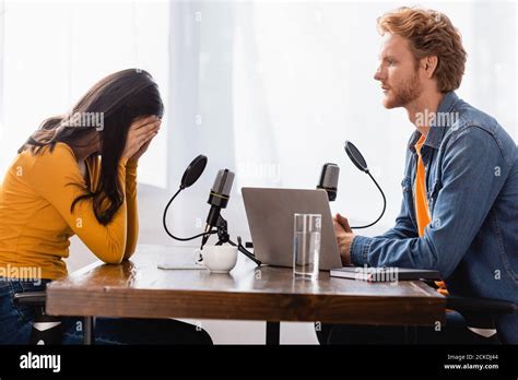 Side View Of Redhead Broadcaster Looking At Stressed Brunette Woman Obscuring Face With Hands