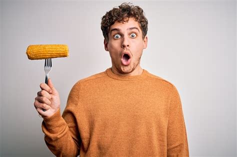 젊은 금발의 잘 생긴 남자 와 곱슬 머리 들고 포크 와 Cob 옥수수 위에 흰색 배경 무서워 충격으로 충격 두려움과 두려움