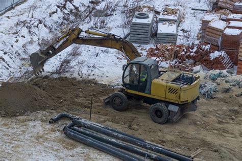 굴삭기는 겨울에 건설 현장에서 트렌치를 파고 있습니다 벽돌 콘크리트 석판 및 건설 폐기물이 건설 현장에 누워 있습니다 노란 굴삭기가 현장 주변에 모래를 던진다 0명에 대한