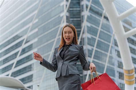 회색 사무실 정장을 입은 젊은 아시아 사업가가 빨간 쇼핑 종이 봉지를 손에 들고 중앙 비즈니스 지구를 행복하게 걷고 있다 18 19세에 대한 스톡 사진 및 기타 이미지