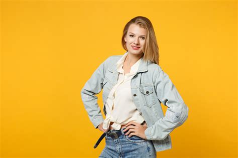 데님 캐주얼에서 아름 다운 젊은 여자의 초상화는 팔에 서 있는 허리에 Akimbo 스튜디오에서 밝은 노란색 오렌지 배경 벽에 고립 사람들 라이프 스타일 개념입니다 복사