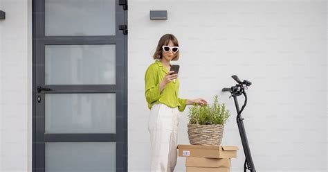 근처에 전기 스쿠터와 소포가 있는 집 현관에 서서 전화를 사용하는 여성 지속 가능성과 현대적인 친환경 라이프 스타일의 개념 사진 Unsplash의 수송 이미지