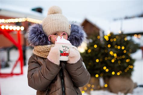 눈 사탕 지팡이 비문 메리와 브라이트가 있는 머그잔을 든 소녀는 축제 시장에서 겨울에 따뜻한 옷을 입고 야외에서 손을 들고 있습니다 요정 조명 화환은 새해를 위해 눈 마을을