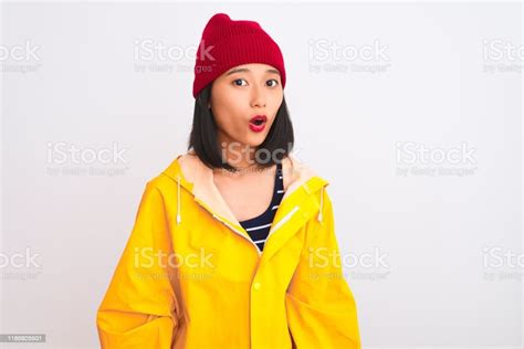 고립 된 흰색 배경에 비옷과 양모 모자를 쓰고 있는 젊은 아름다운 중국 여성은 놀라움과 두려움 흥분된 얼굴로 충격을 받았습니다 가을에 대한 스톡 사진 및 기타 이미지