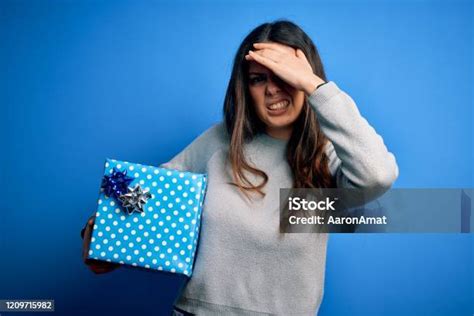 고립 된 파란색 배경 위에 생일 선물을 들고 젊은 아름다운 갈색 머리 여자 부끄러움과 깜짝 얼굴 분노와 좌절에 충격 머리에 손으로 스트레스 실수에 대한 두려움과 화가 갈색