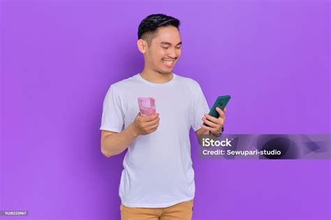 스마트 폰을 사용하여 흰색 티셔츠를 입고 보라색 배경에 고립 된 돈 지폐를 들고있는 쾌활한 젊은 아시아 남자 30 34세에 대한 스톡 사진 및 기타 이미지 Istock