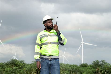 안전 조끼와 헬멧을 쓴 아프리카 엔지니어가 비가 내린 후 풍력 터빈 농장에서 무전기를 사용하고 있습니다 노동자는 하늘에 무지개가 있는 풍차 공원에서 일합니다 공학 생태학