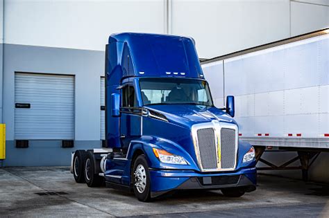 블루 보닛 데이 캡 큰 장비 세미 트럭 트랙터 지붕 스포일러가 창고 도크 게이트 드라이 밴 세미 트레일러에 적재 옆에 서서 다음화물을 준비합니다 차량 트레일러에 대한 스톡